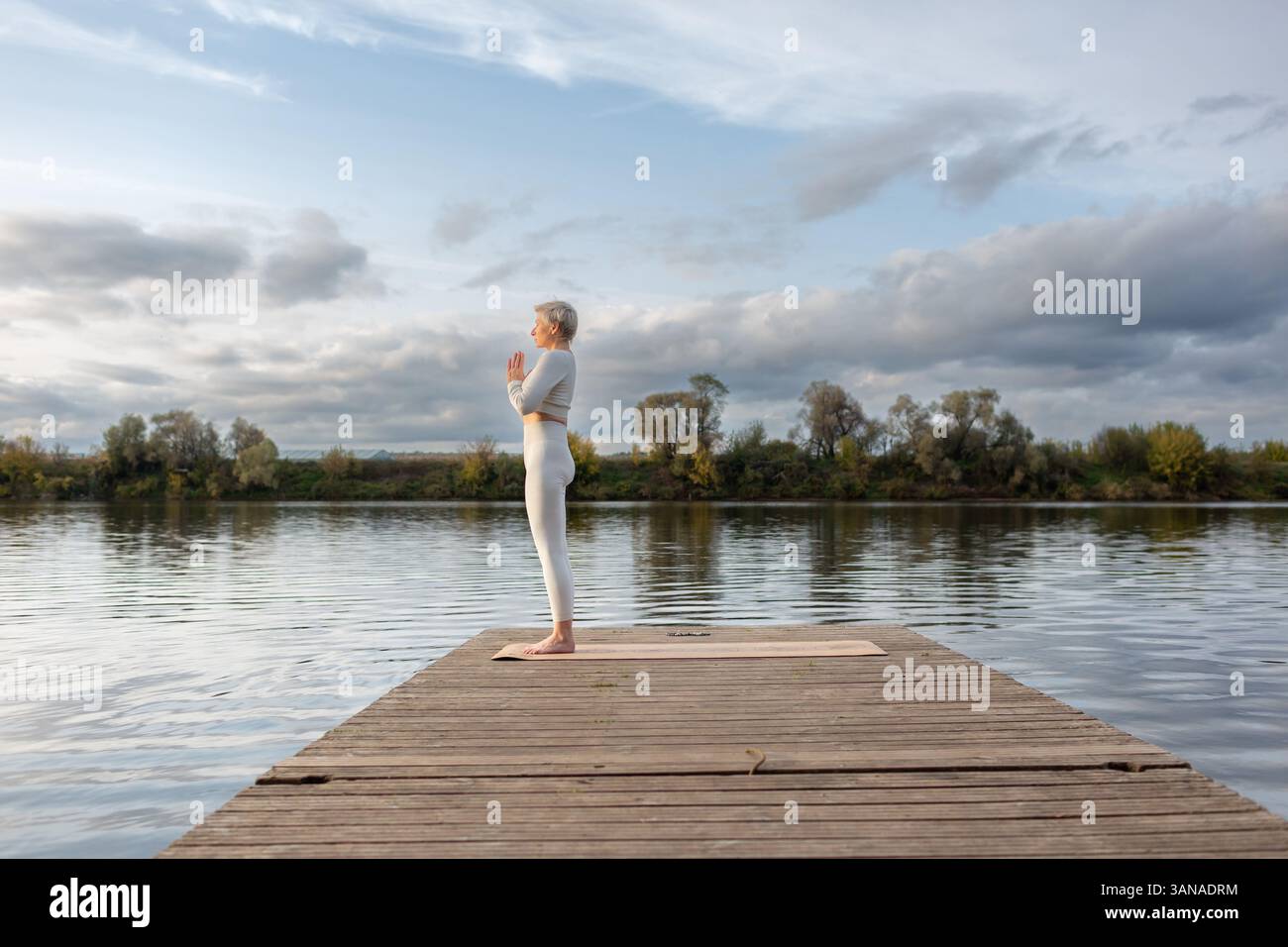 An adult woman in a white suit performs yoga asanas on a pier by the ...