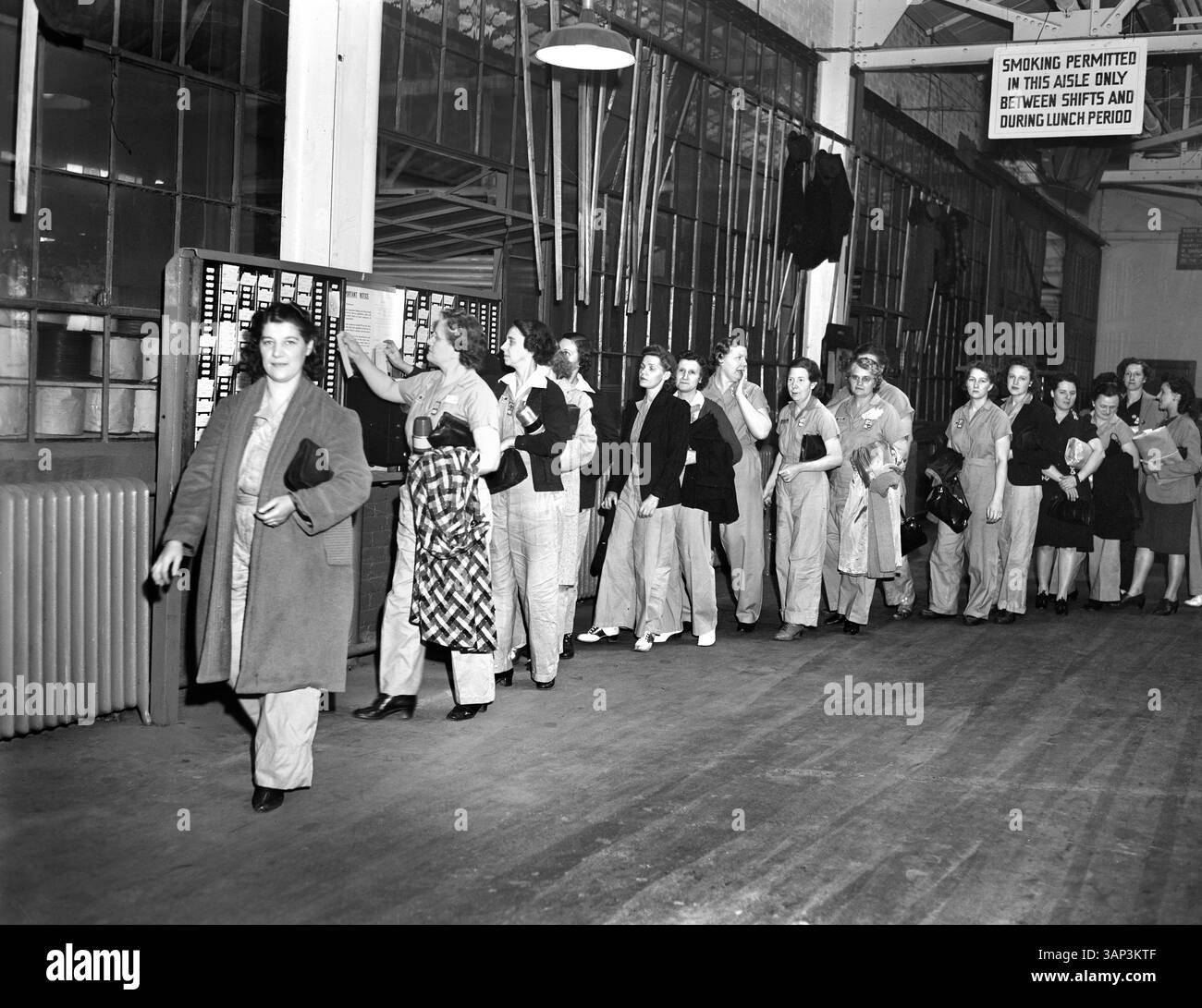 Women war workers wearing slacks punching time cards at end of shift ...