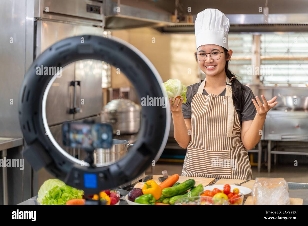 A young female chef wearing a striped apron and white chef hat records ...