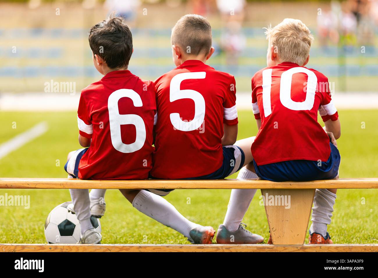 Boys in red soccer jerseys with white numbers sit on a wooden ...
