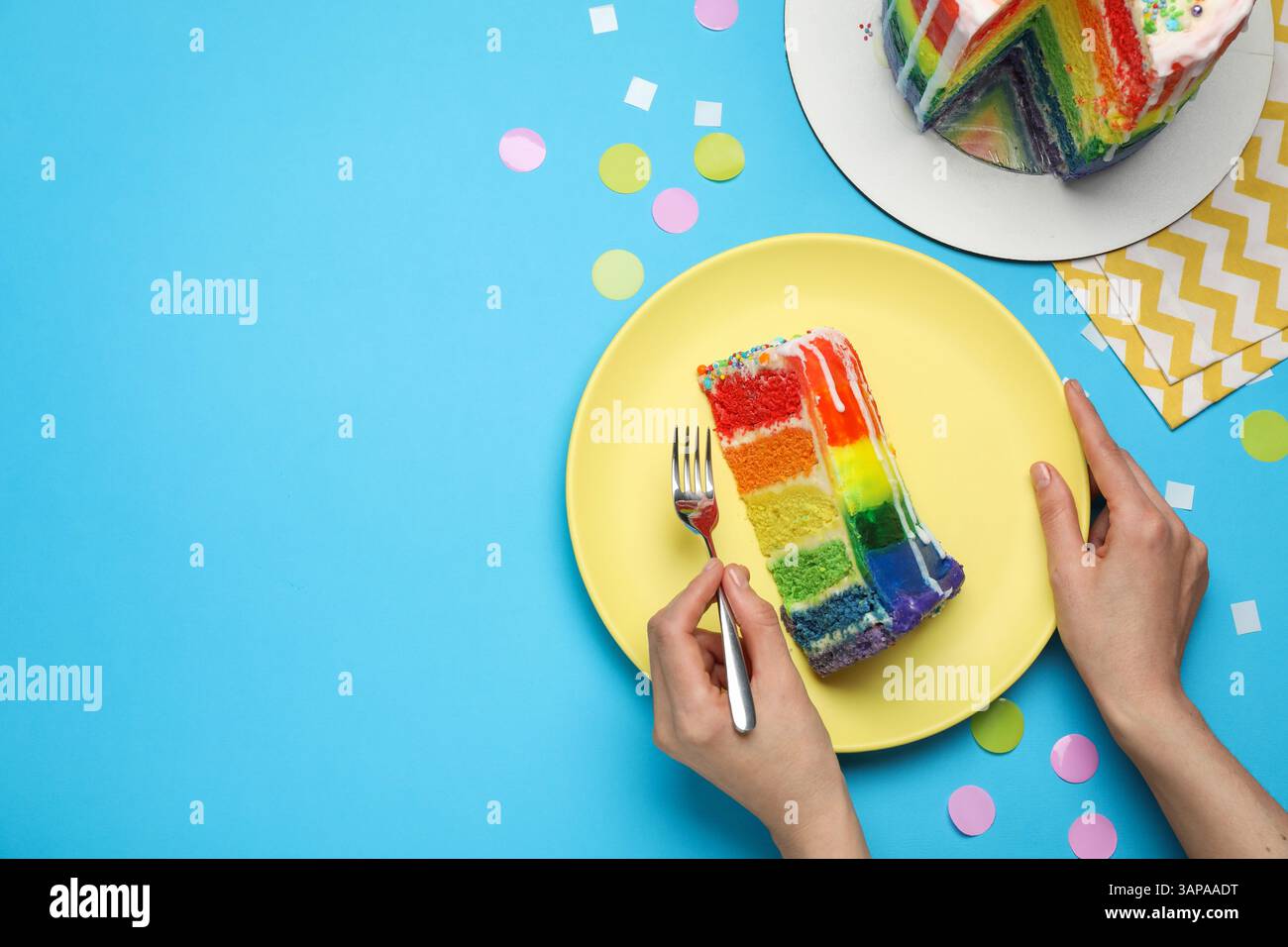 Woman eating delicious rainbow cake at light blue table with confetti ...