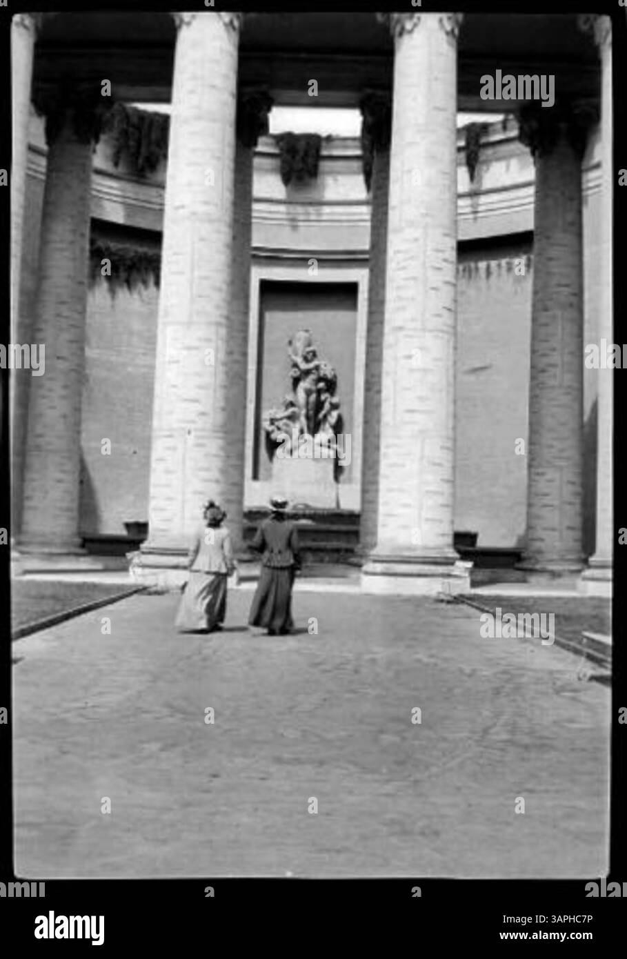 Two women in long skirts and hats stroll down a walkway, passing a ...