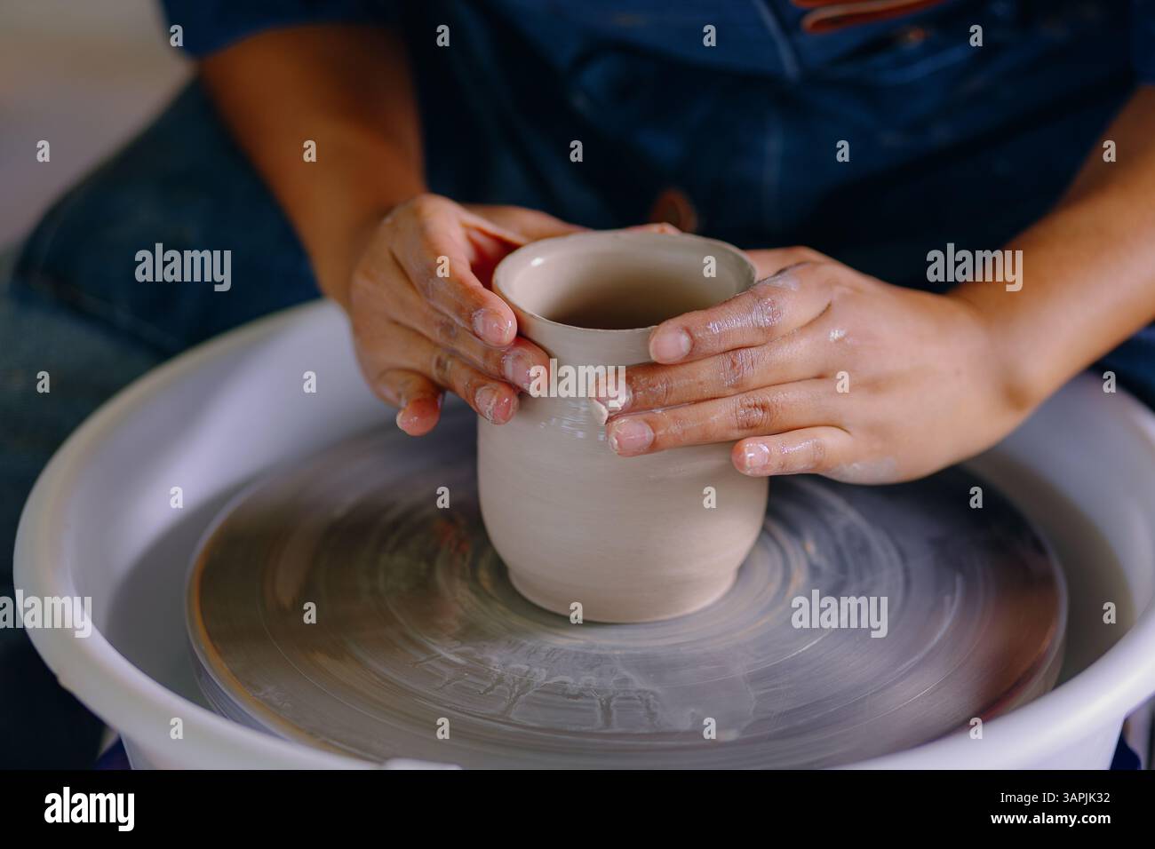Mexican woman, 24, molds a ceramic piece in her studio, wearing a blue apron and denim. Close-up ...