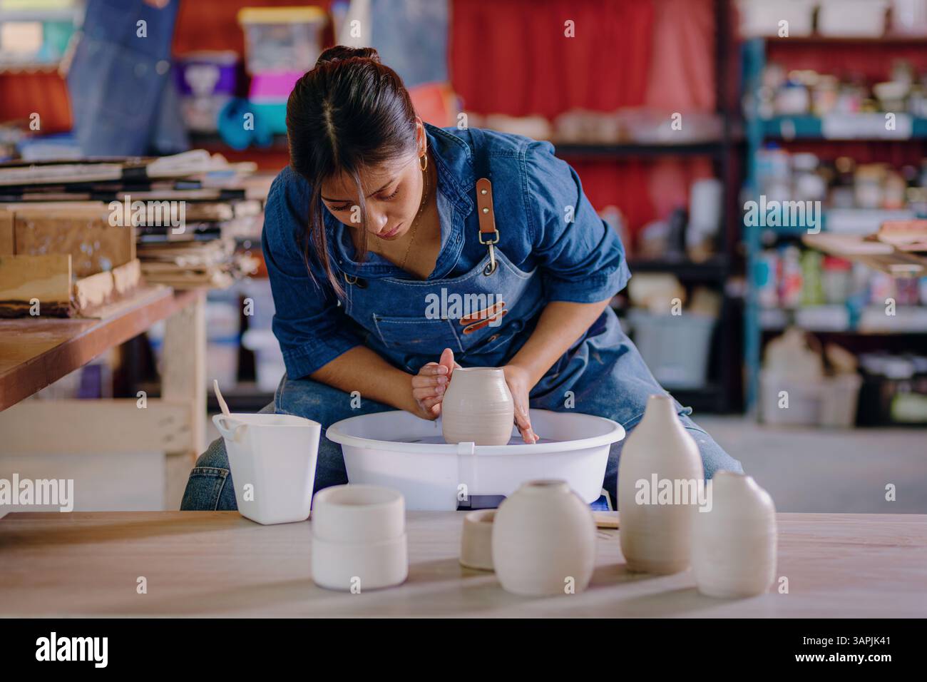 Mexican woman, 24, molds a ceramic piece in her studio wearing a blue apron, shirt, and jeans ...