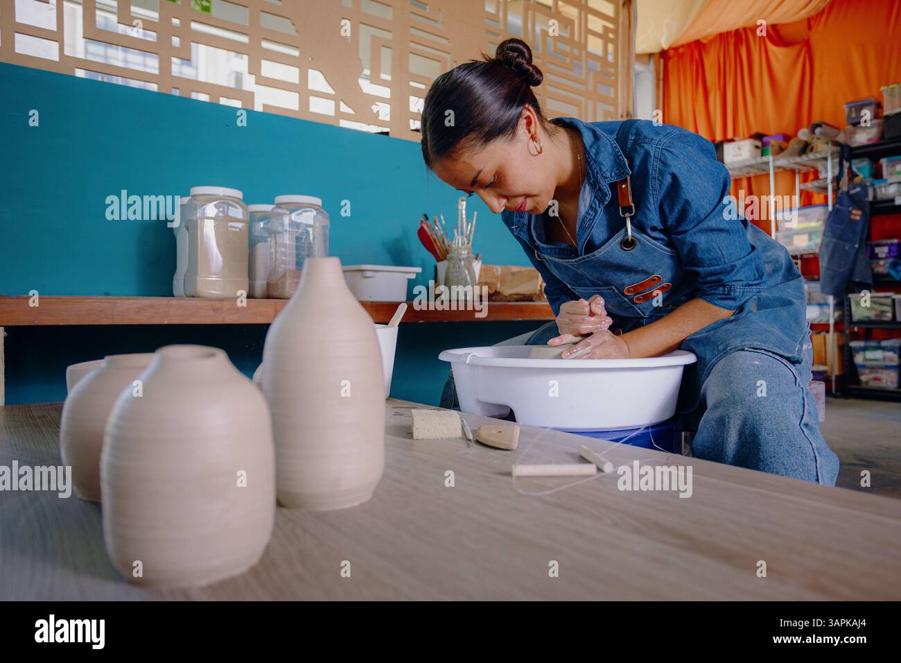 Mexican woman, 24, molds a ceramic piece in her studio wearing a blue apron, shirt, and jeans ...
