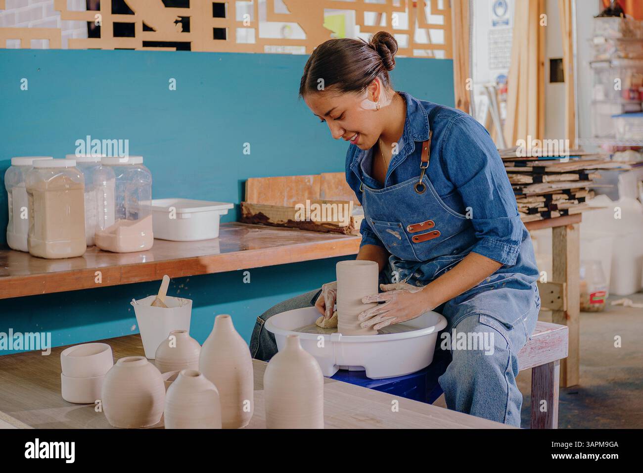 Mexican woman, 24, molds a ceramic piece in her studio wearing a blue apron, shirt, and jeans ...