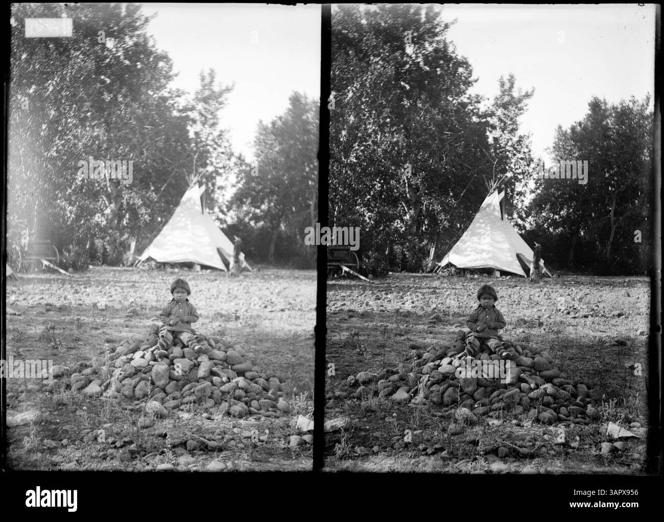 Photograph of a child seated on a pile of rocks in front of a tipi. The ...