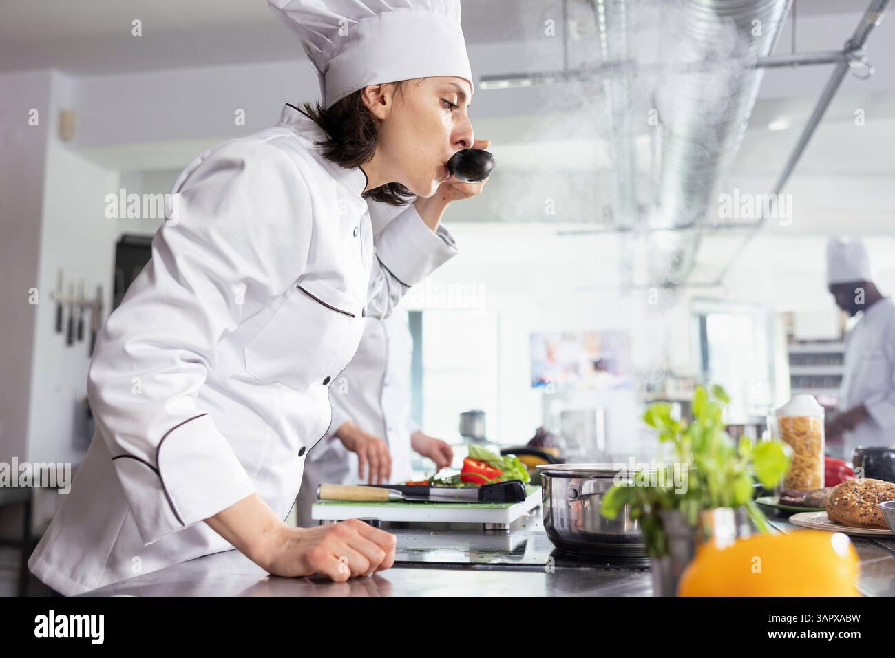 Female chef in uniform leans over the stovetop and tasting a delicious ...