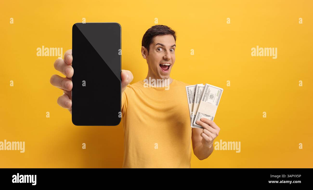 Happy young man holding money and smartphone against yellow background ...