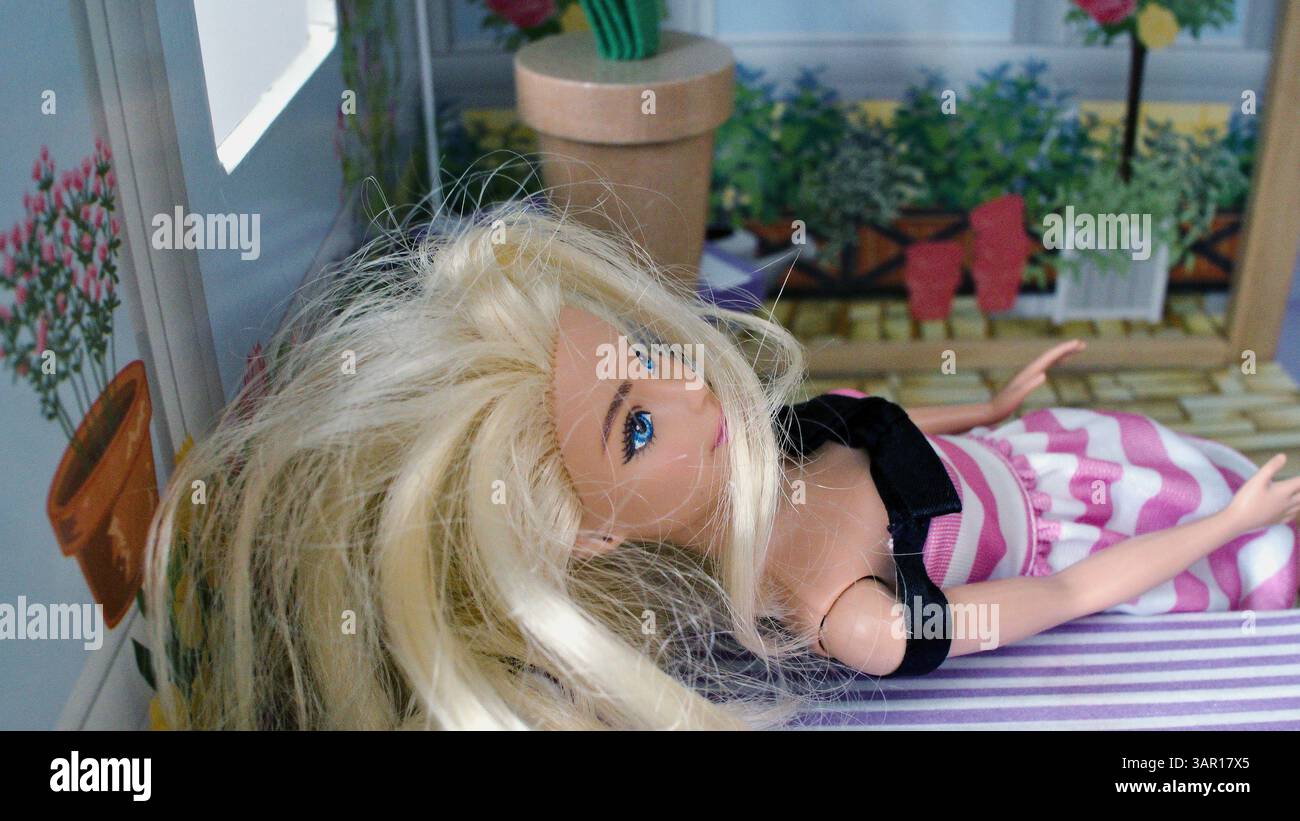 A close-up of a blonde doll lying on a striped bed in a miniature ...