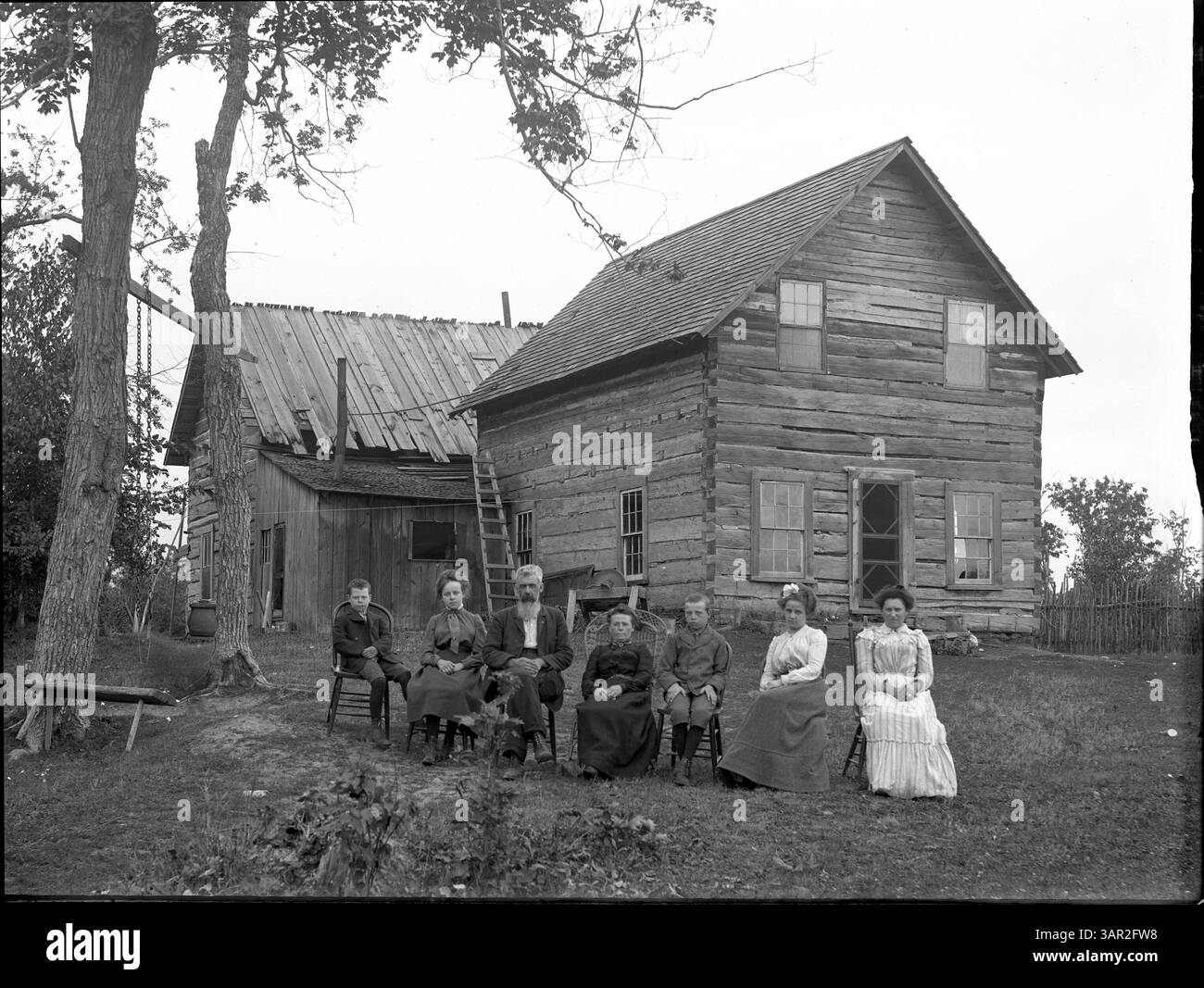 The photograph shows a family outside a log house with an attached barn ...