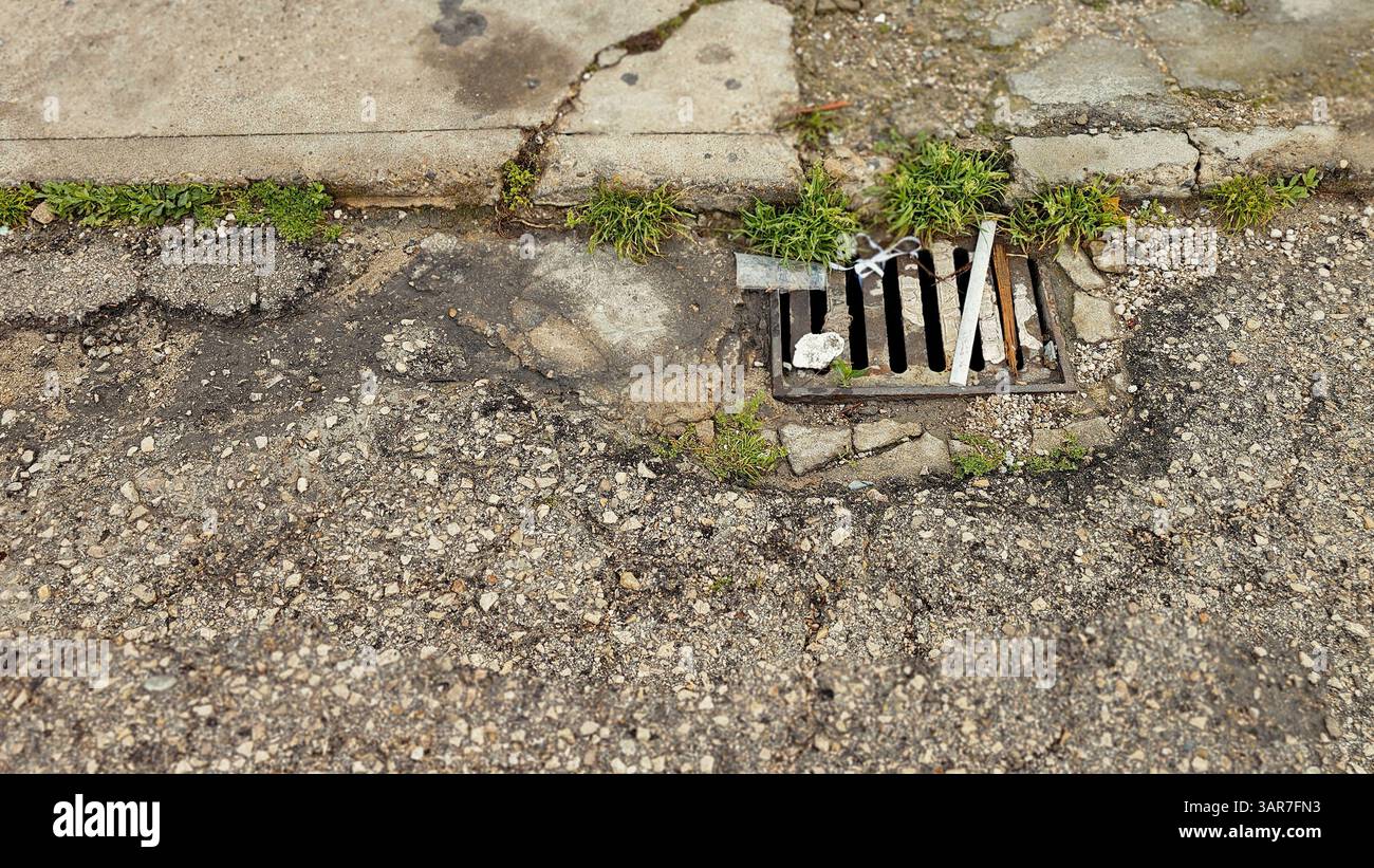 Damaged asphalt road with a sewer grate, weeds, and debris, showing ...
