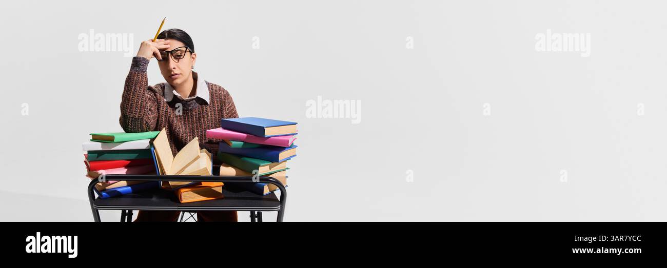 Exhausted teacher sits surrounded by messy stacks of books, holding ...