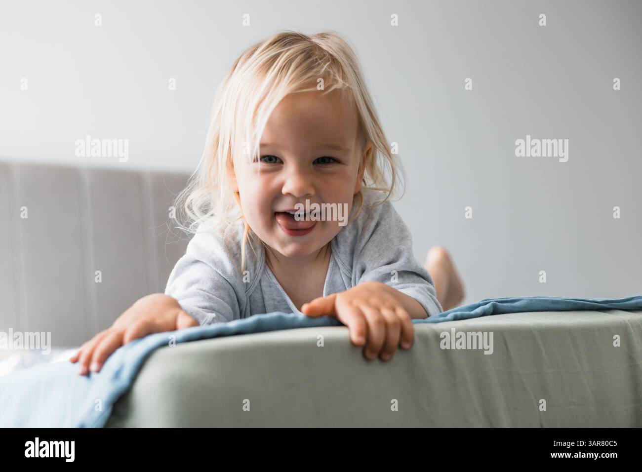 A happy blonde girl lies on her stomach in bed, smiling widely. The ...
