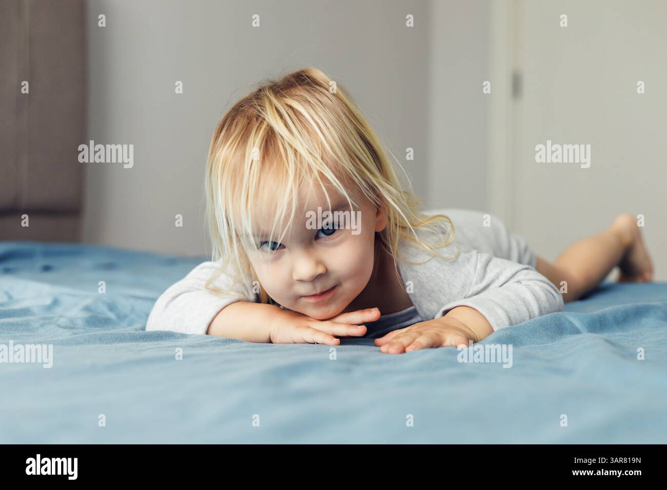 A beautiful blonde toddler girl is playfully lying on a bed at home ...