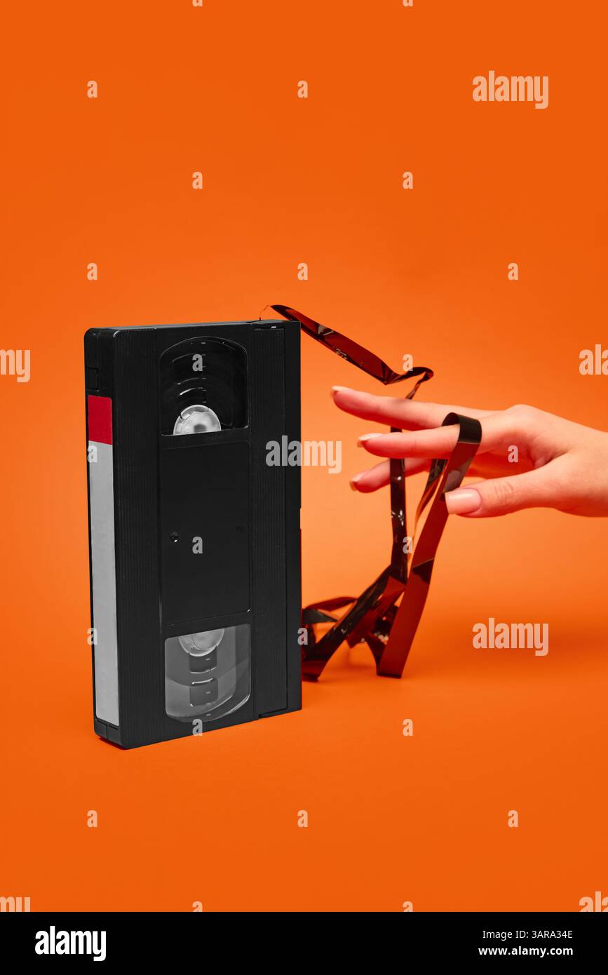 Hand with tangled VHS tape extending from cassette on orange background ...