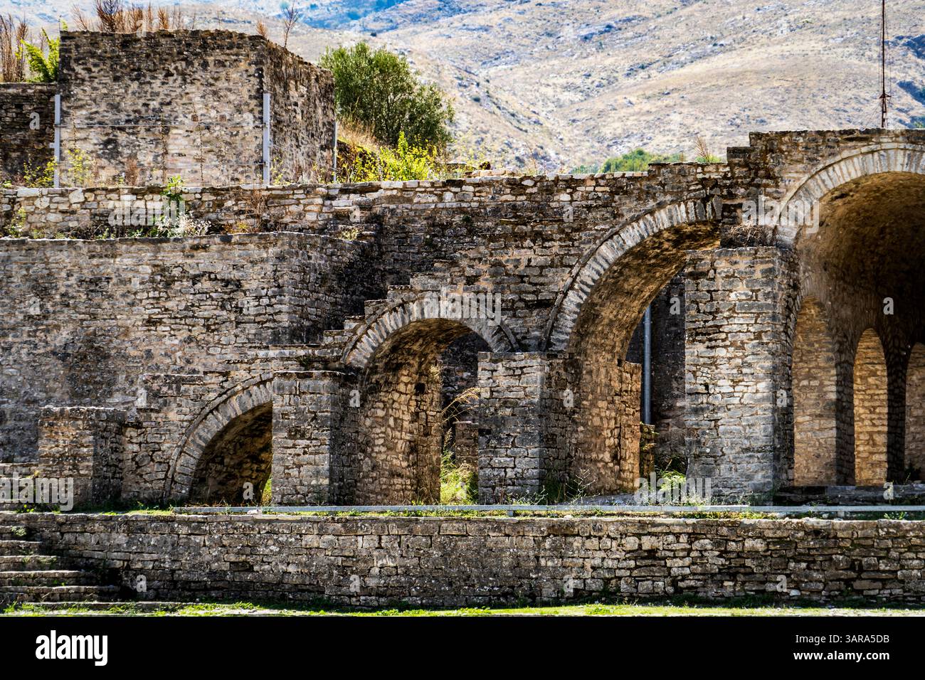 Ancient stone ruins featuring arches and walls, surrounded by greenery and mountains in the ...