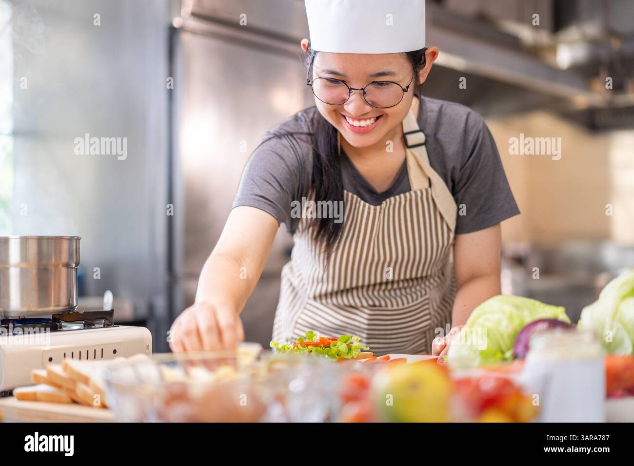 A cheerful young female chef wearing a striped apron and white chef hat ...