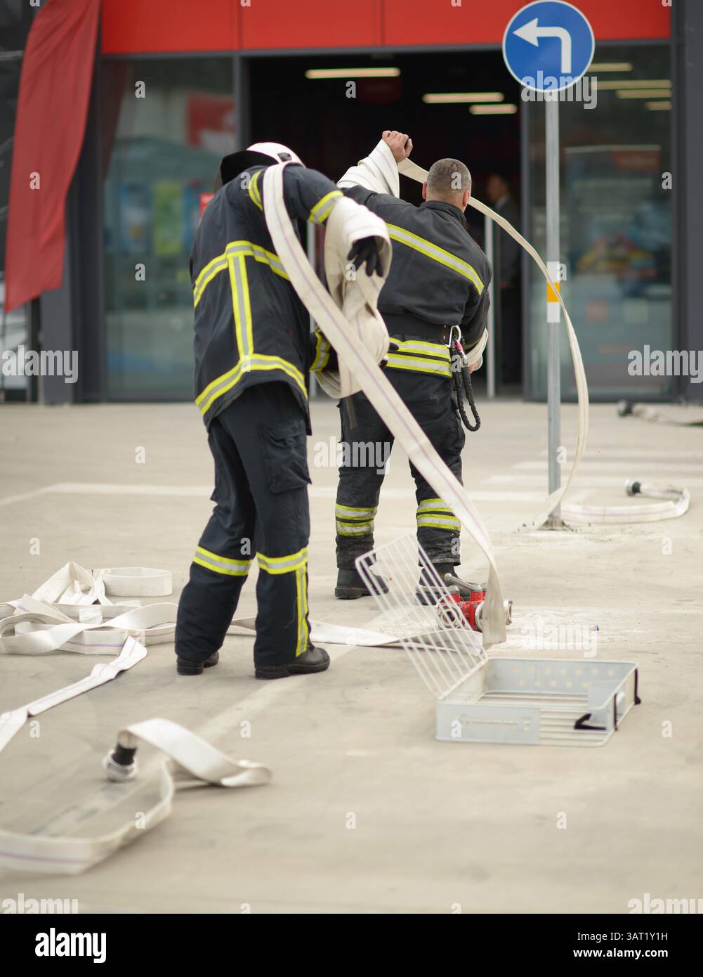 Two firefighters in full protective gear unroll a fire hose on a paved ...