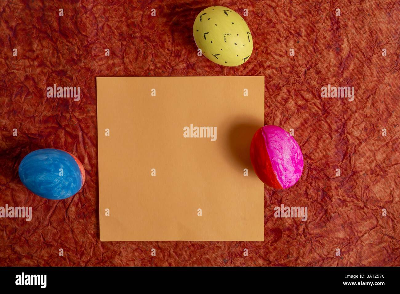 Brightly colored Easter eggs, placed on a rough orange surface around a plain orange paper ...