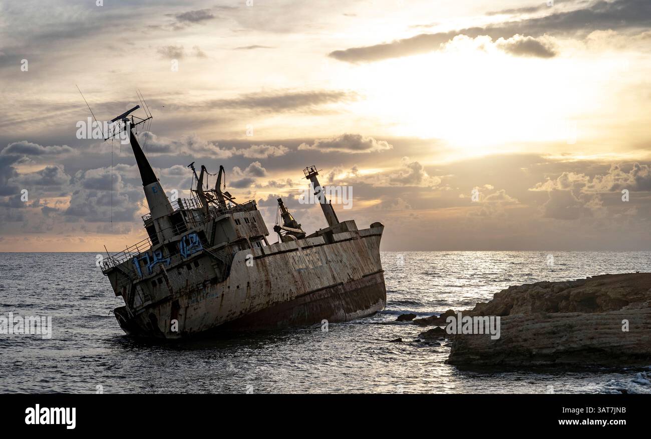 A rusted shipwreck leaning on rocks at sea during a dramatic sunset ...