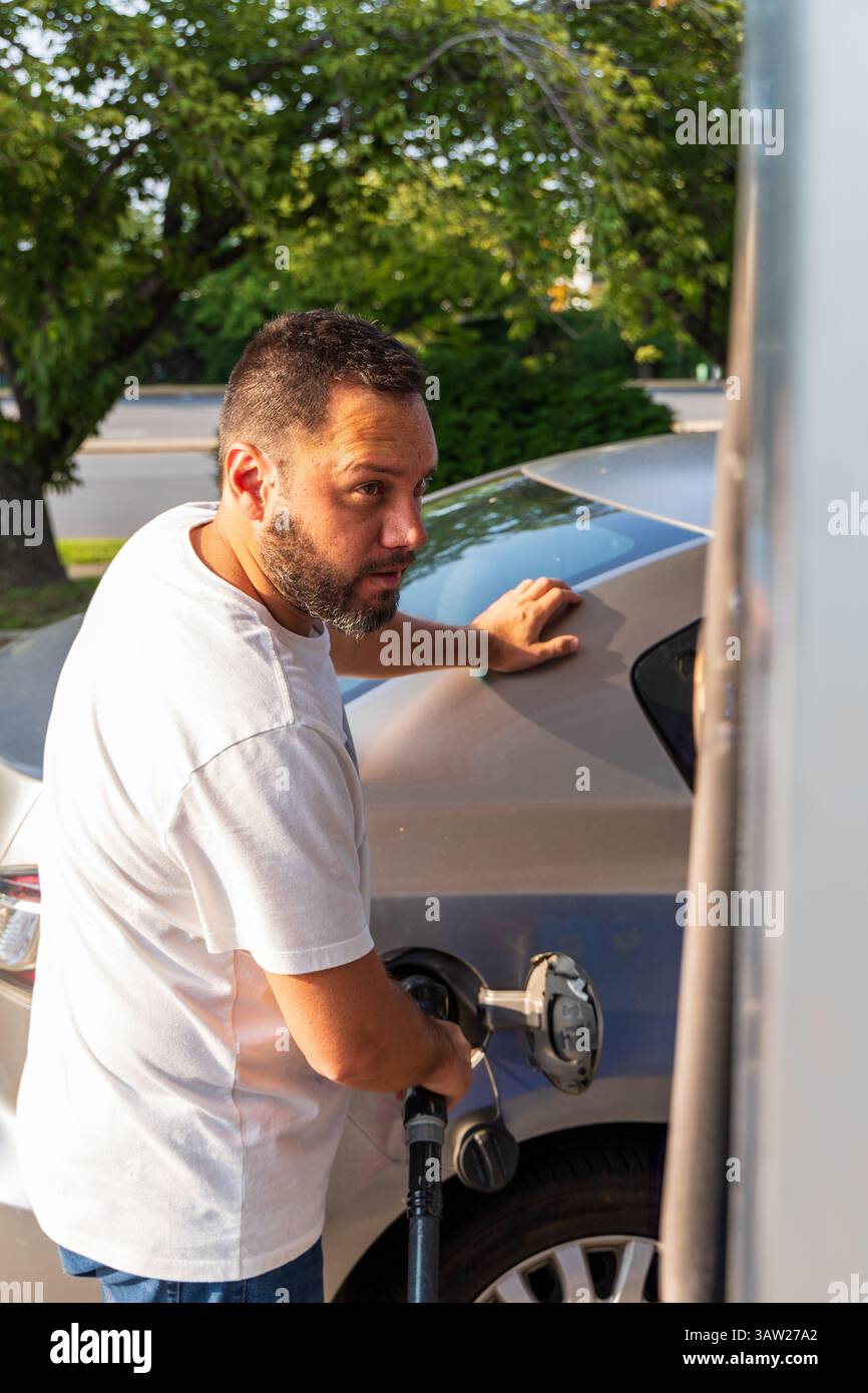 A man is filling up his car at a gas station. He is wearing a white ...