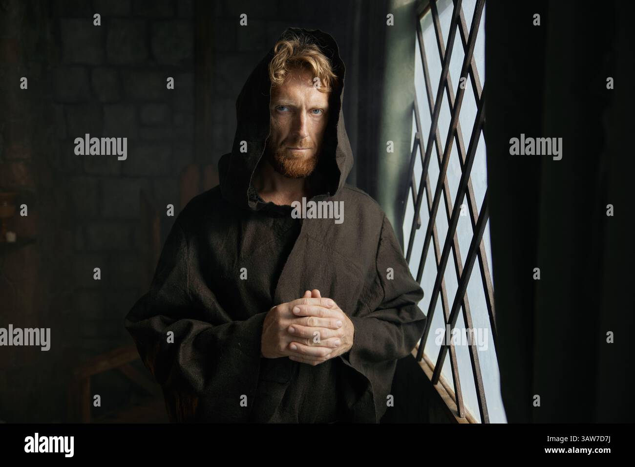 Pensive monk in medieval costume with hood looking through window of ...