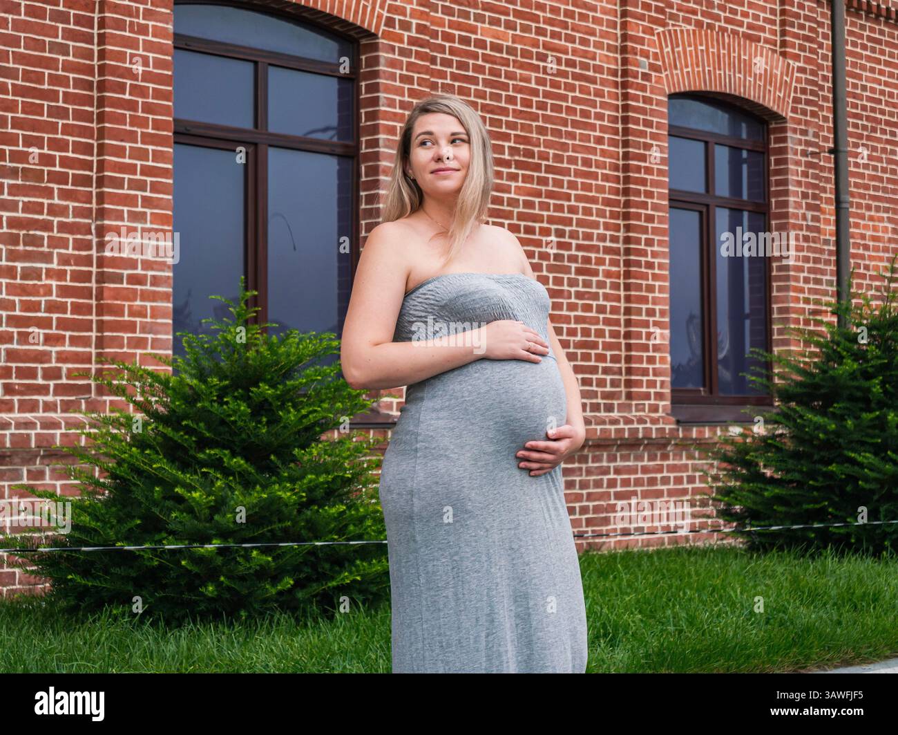 Pregnant woman cradles her baby bump while standing outdoors by a brick ...