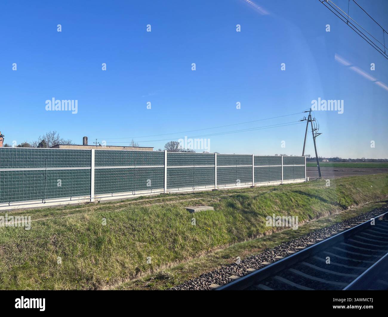 A rural railway landscape with a metal noise barrier fence running parallel to the train tracks ...