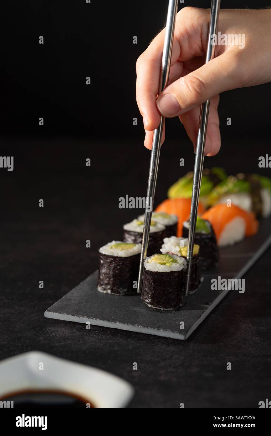 A hand taking a piece of avocado maki with stainless steel chopsticks from a sushi plate. It ...