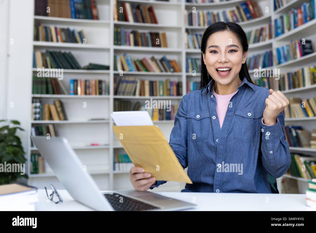 An excited asian woman holds a letter, celebrating success in a library ...