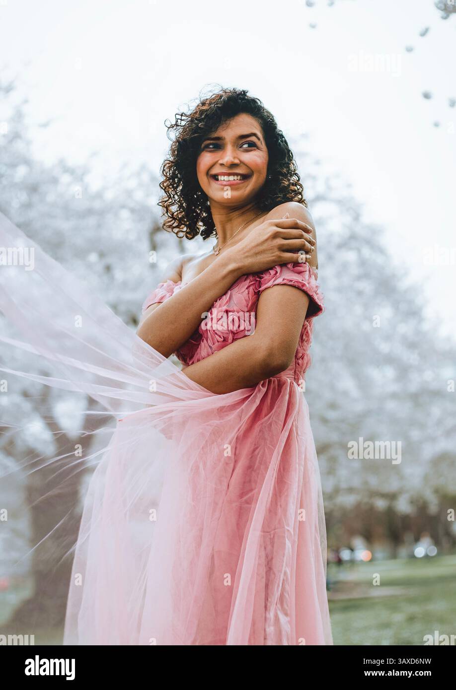 A woman smiles wrapping her arms around herself in a long pink dress ...