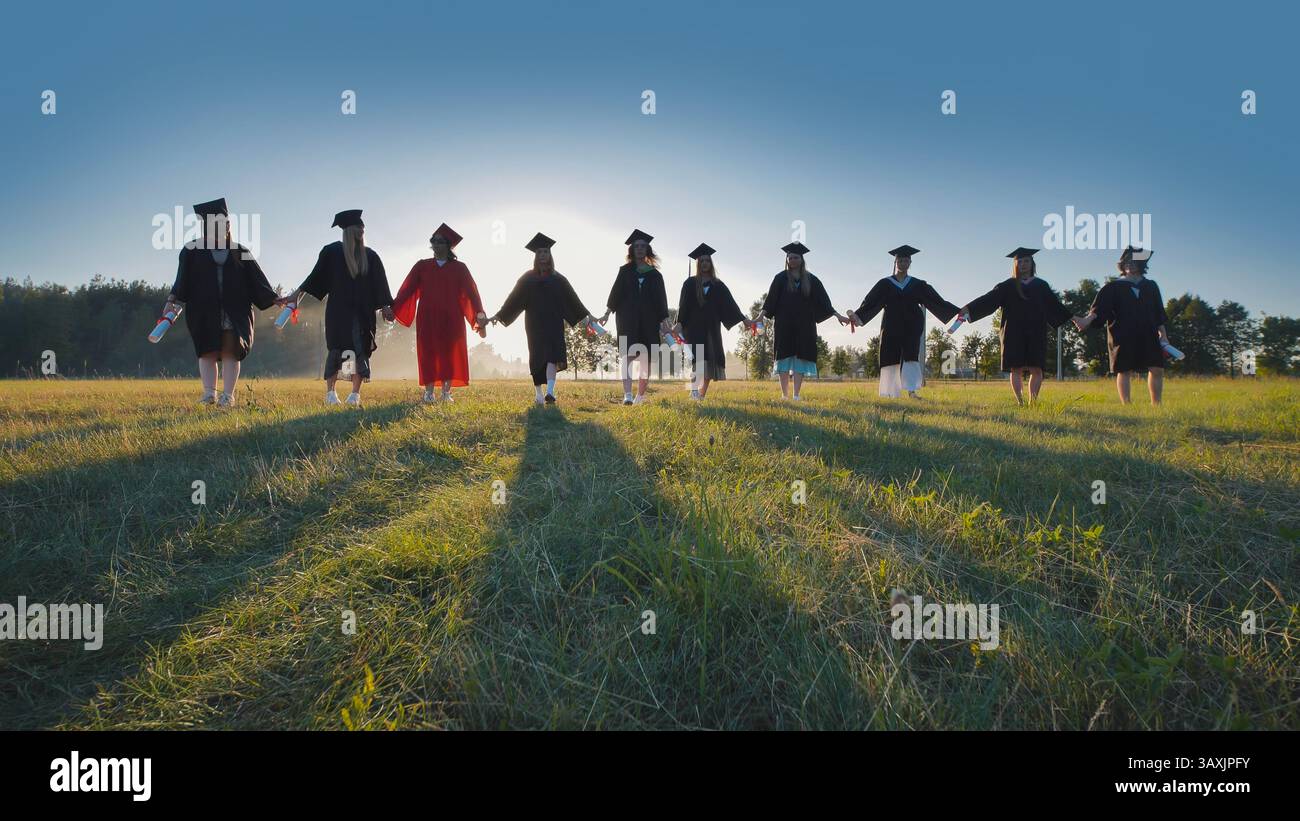 Group of graduates is walking in a field at sunset, holding hands and ...