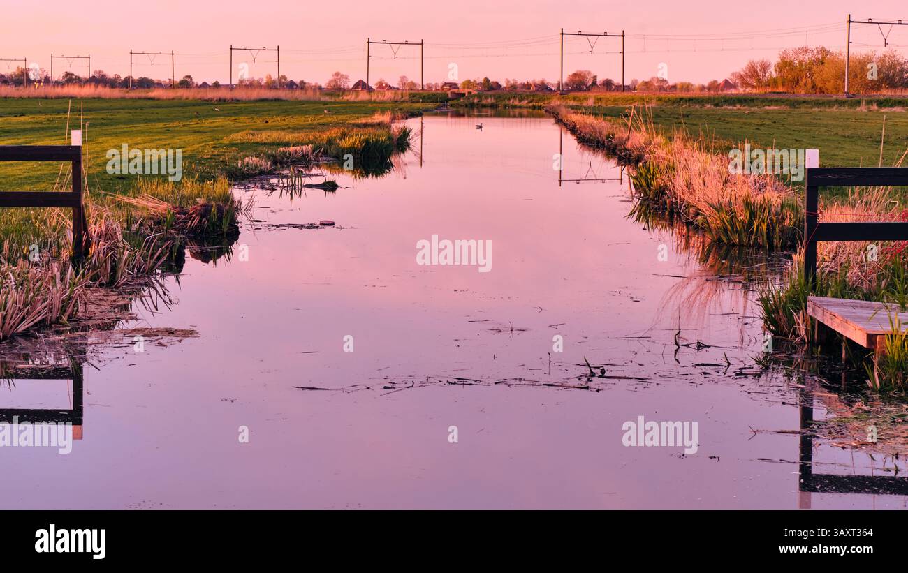 Wide canal in a typical Dutch meadow bird reserve at sunset, with railway pylons crossing the ...