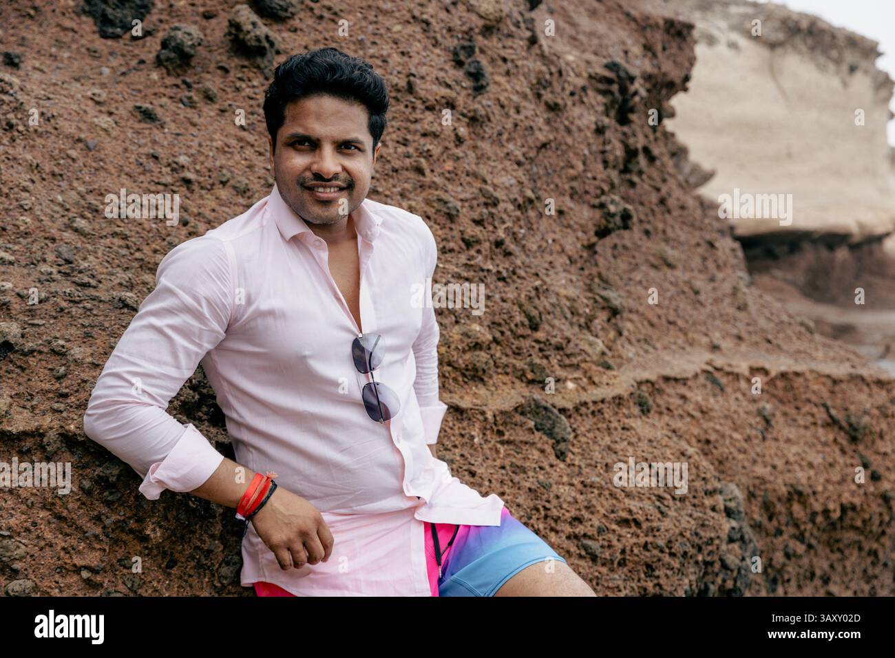 A confident smiling Indian man posing outdoor leaning on a rock at the ...