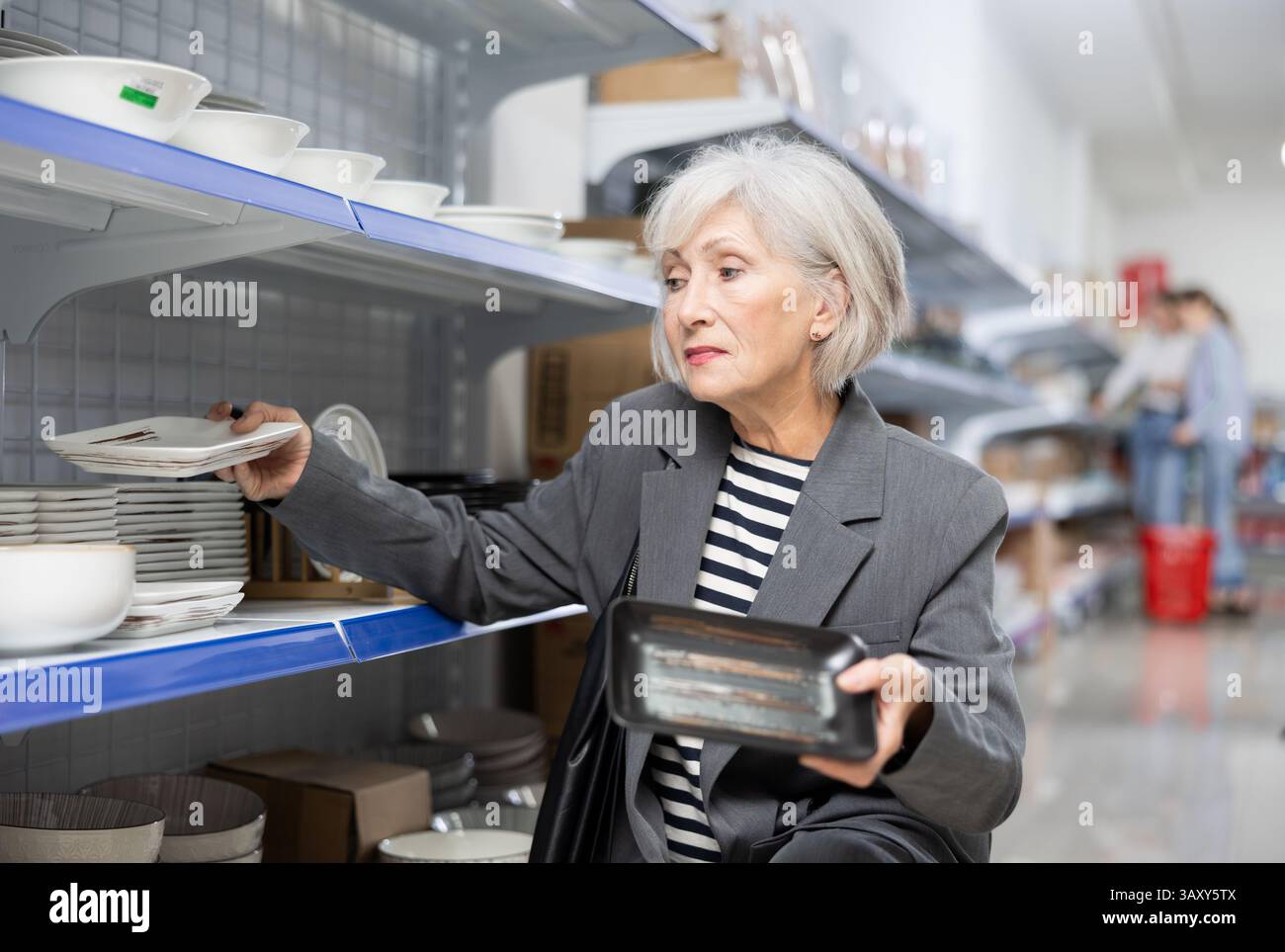 Enthusiastic senior lady choosing square ceramic plates at Asian store ...
