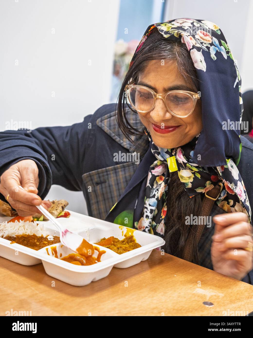 An Indian woman wearing glasses & a floral headscarf sits at a table ...