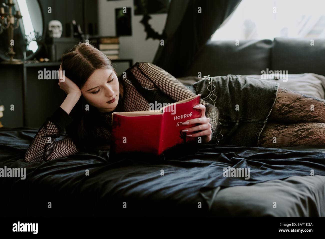 Goth teen resting on double bed with black linen while reading horror ...