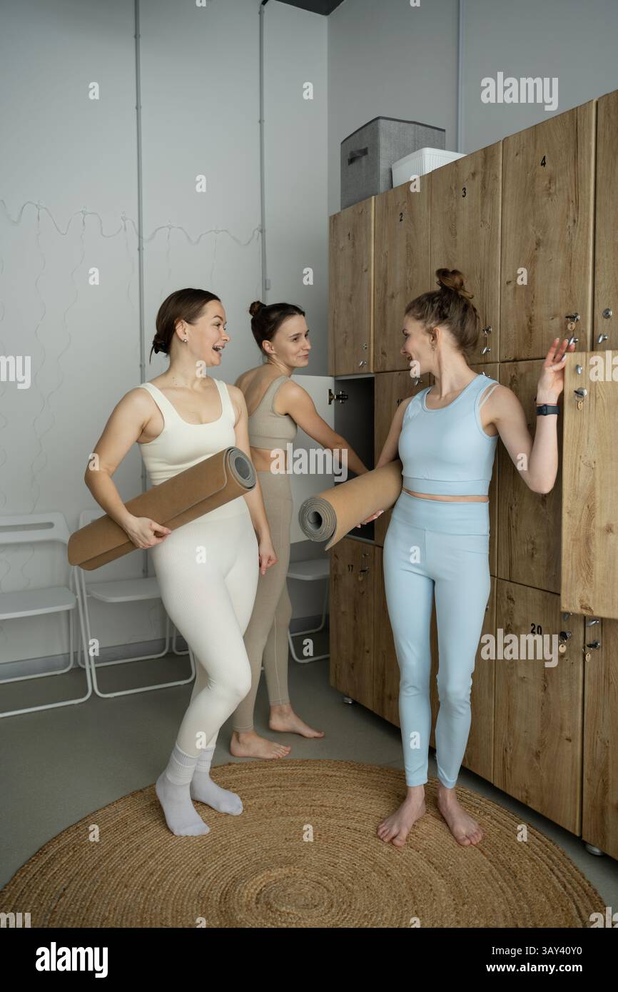 Three women in active wear chat animatedly in a modern locker room before a yoga class, holding ...