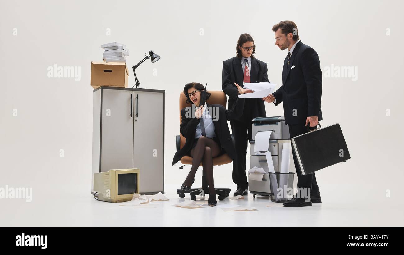 Three office workers gathered around printer, woman yelling on phone ...
