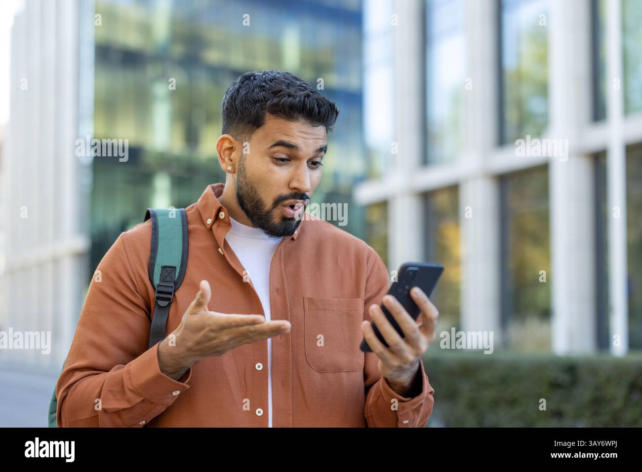 A young man outside reacts with surprise while looking at his smartphone. He gestures with his ...