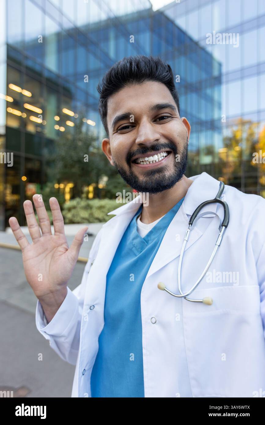 A smiling doctor waves at the camera outside a modern building, wearing ...