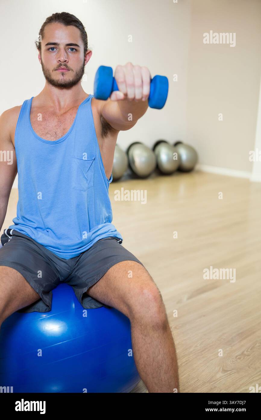 young adult man sitting on stability ball in gym studio, lifting blue ...