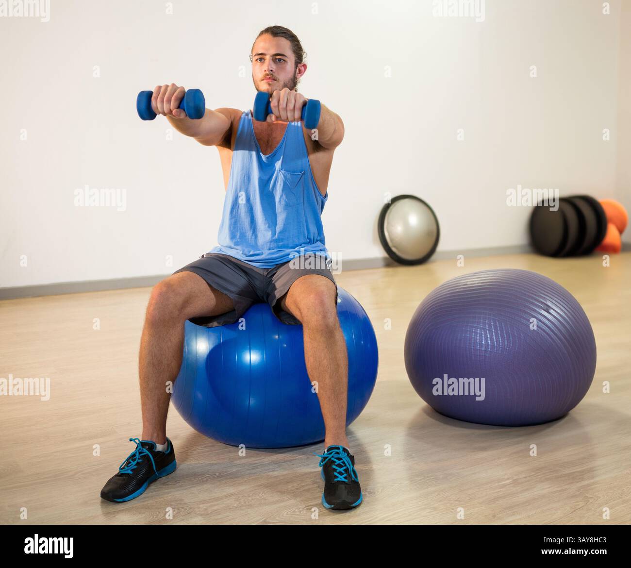 Man sitting on blue stability ball in gym, lifting blue dumbbells with ...