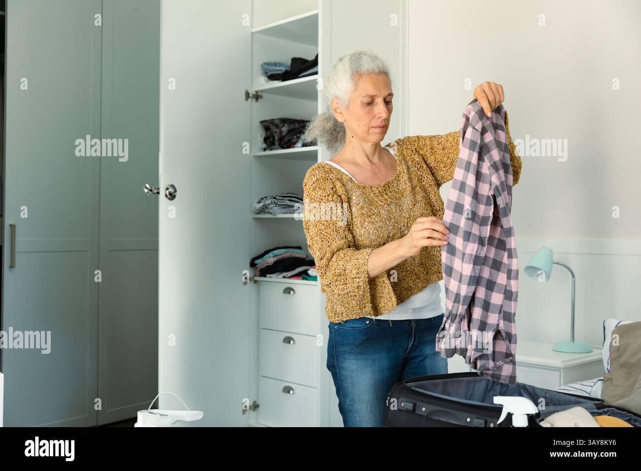 Senior woman sorting clothes from black suitcase in minimalist bedroom ...