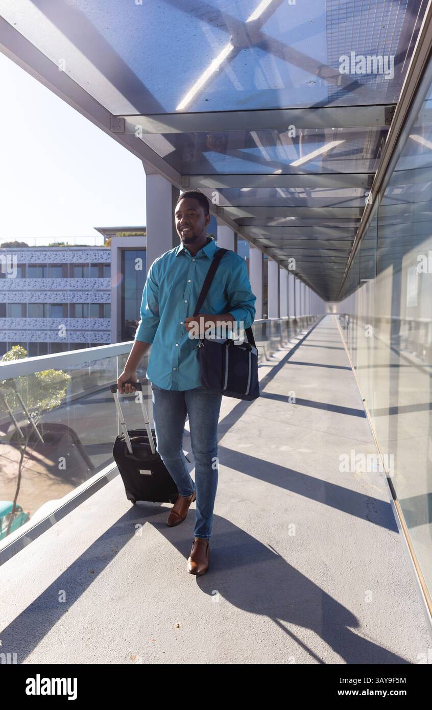 African American man walking glass skybridge, pulling black rolling ...