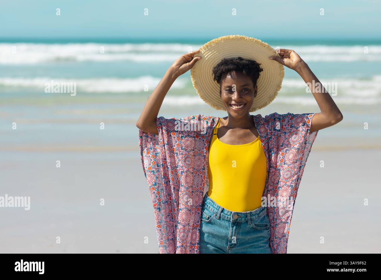 African American woman lifting straw sun hat over head on beach ...