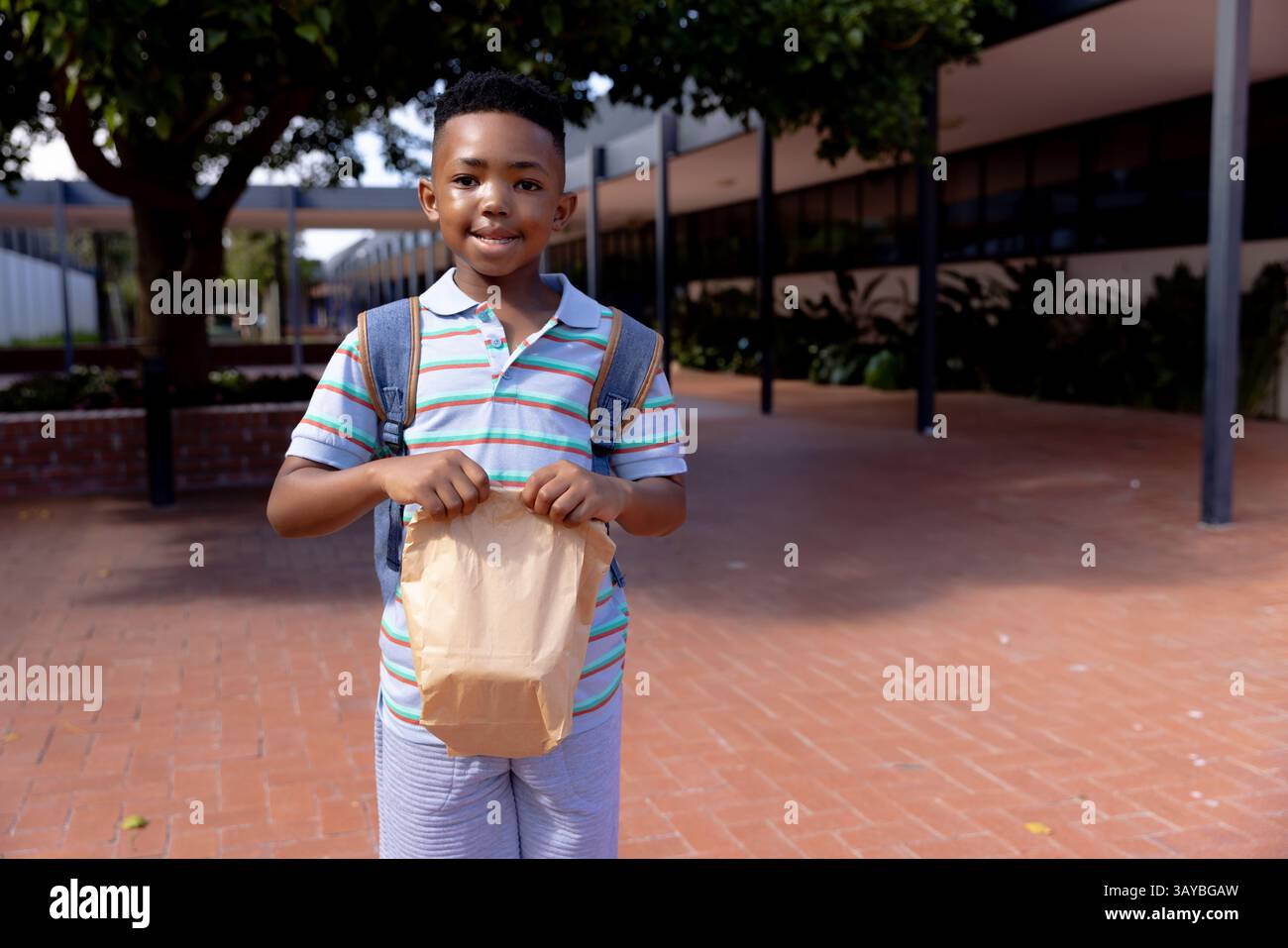 African American boy holding brown paper bag and wearing blue backpack ...