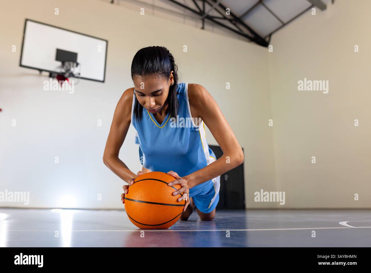 Diverse female athlete kneeling on gym floor pressing orange basketball ...