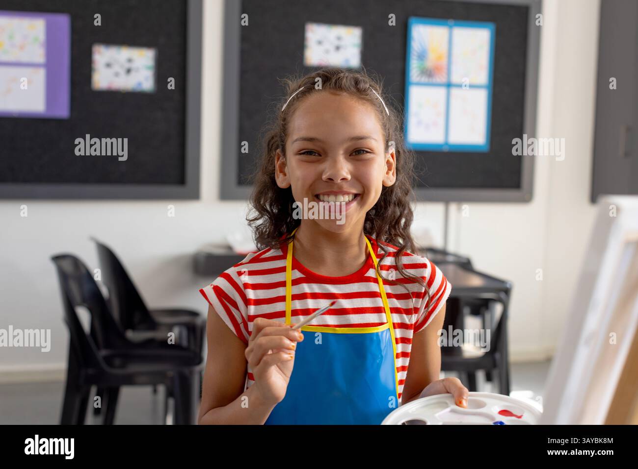 Multiracial school-aged girl painting in school art classroom, holding ...